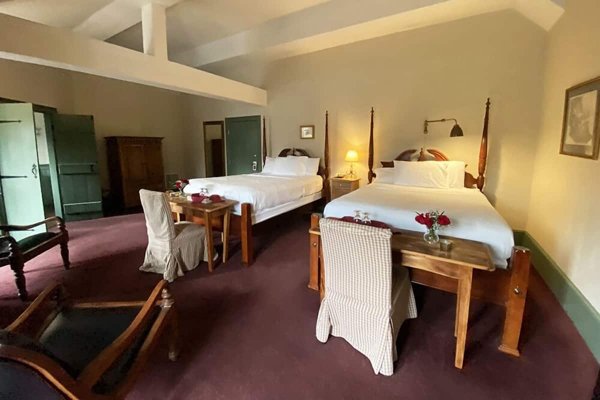 Quarto de hotel com duas camas, uma cadeira de madeira, uma mesa pequena e uma escrivaninha.