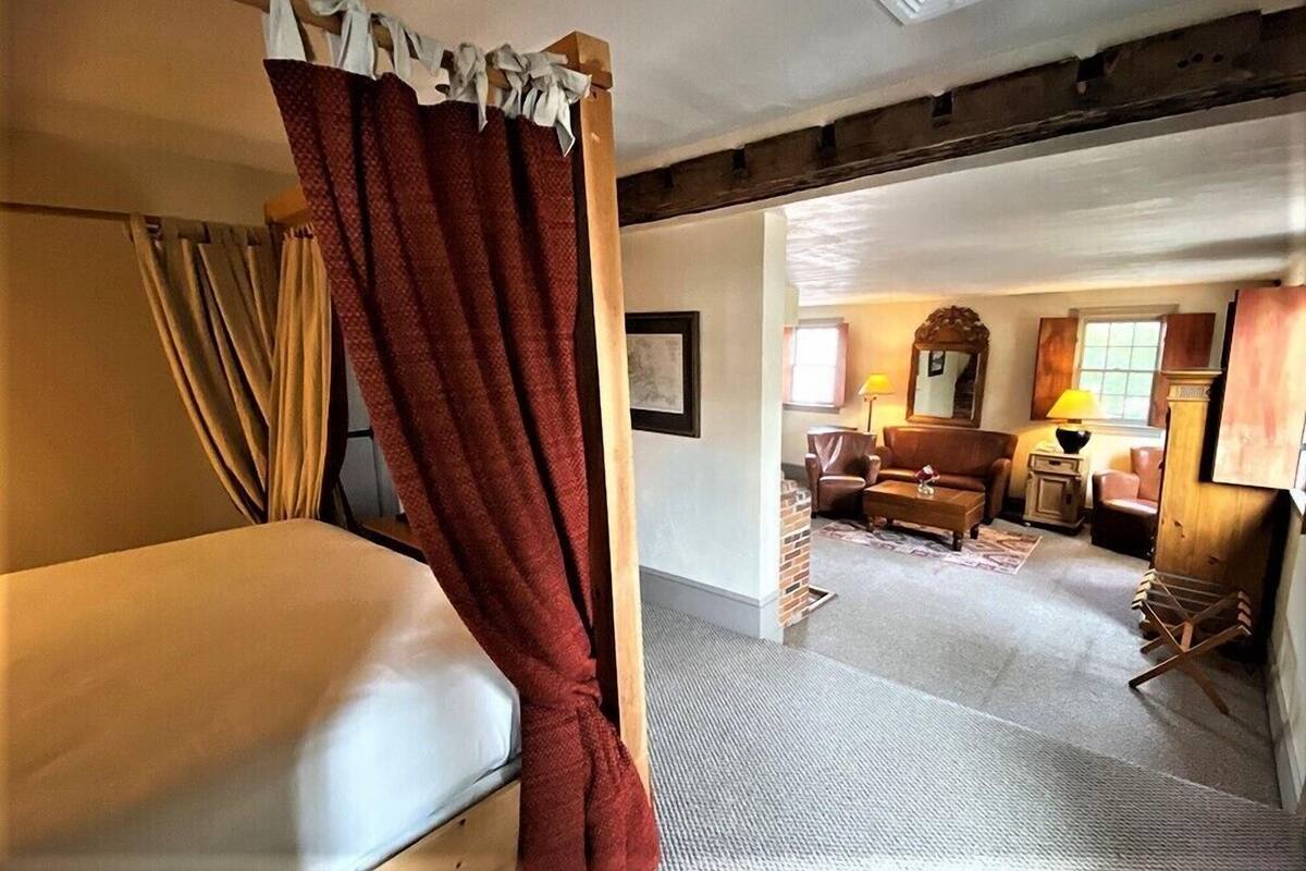 Quarto com cama, cabeceira de madeira e cortina vermelha. Há um corredor que leva a uma sala de estar com sofá, mesa de centro e abajur.
