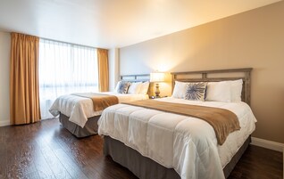 Quarto de hotel com duas camas, cabeceira de madeira, luminária de cabeceira e uma janela grande com cortinas.