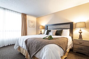 Quarto de hotel com uma cama grande, duas mesas de cabeceira, um criado-mudo, uma luminária e uma janela com cortinas.