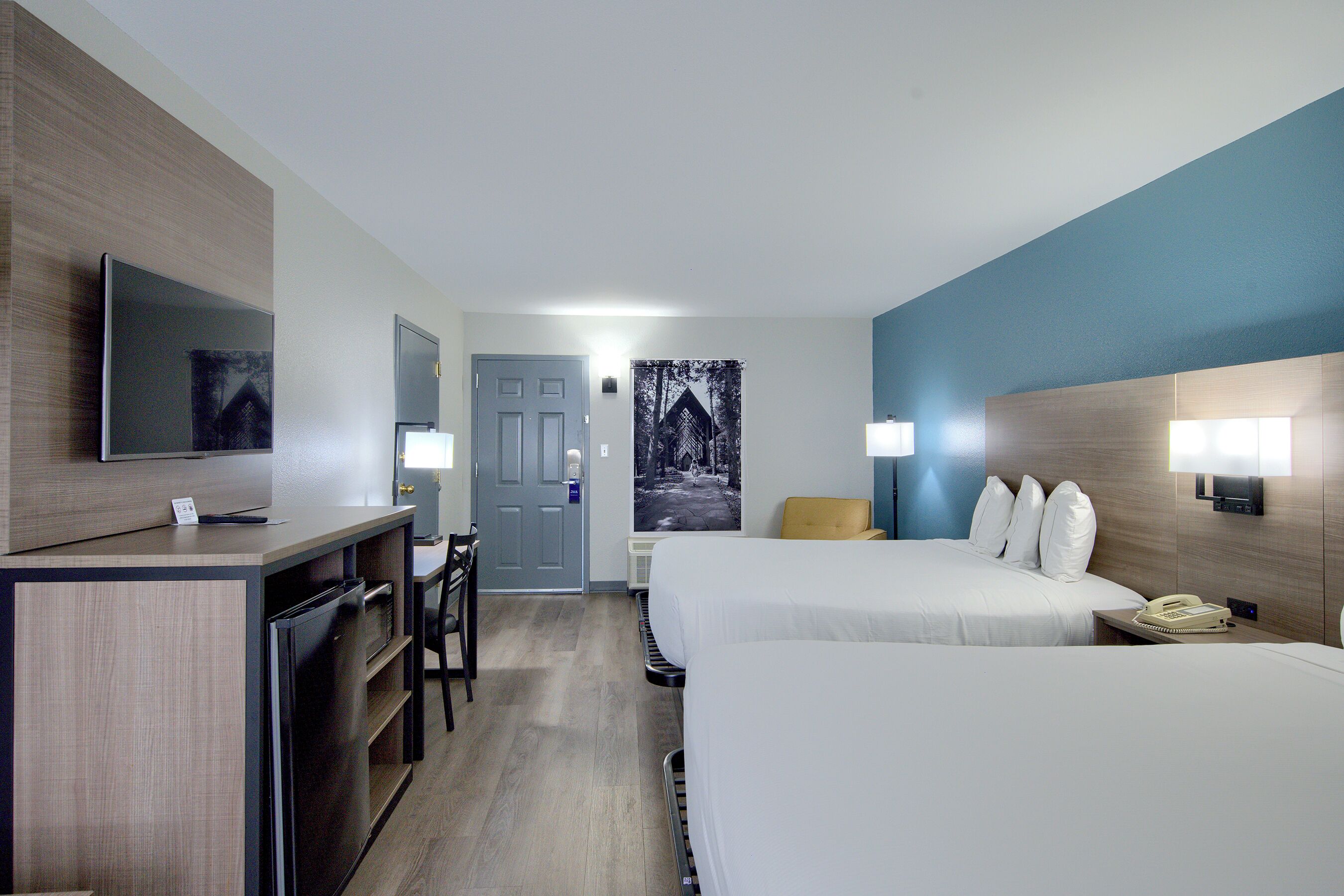 Quarto de hotel com uma cama, escrivaninha com computador, cadeira, porta e telefone fixado na parede.