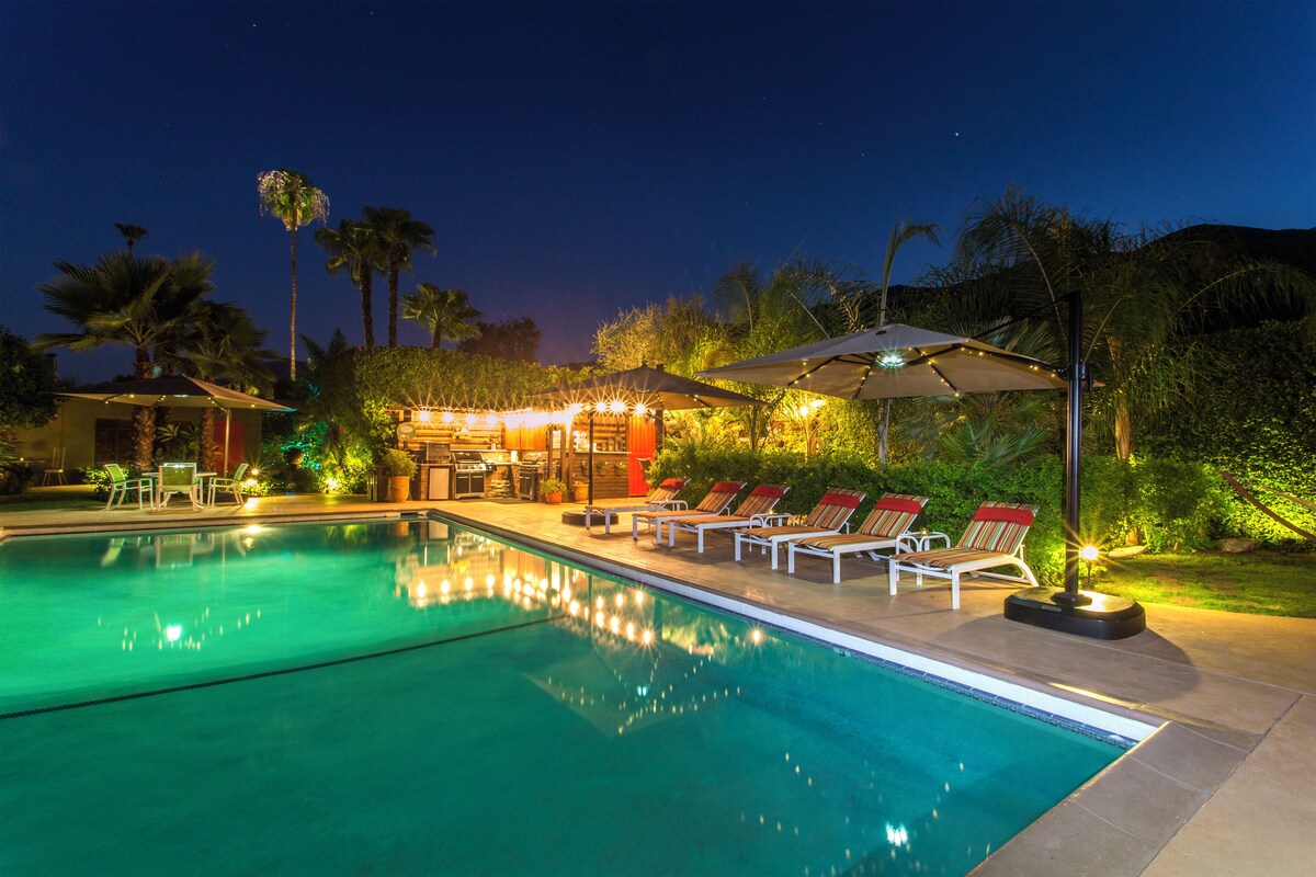 Piscina com espreguiçadeiras e guarda-sóis, rodeada por vegetação e um espaço de bar.