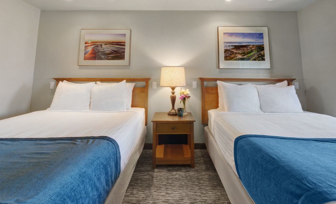 Quarto de hotel com duas camas, uma mesa de cabeceira com luminária e vaso, e duas fotografias emolduradas na parede.