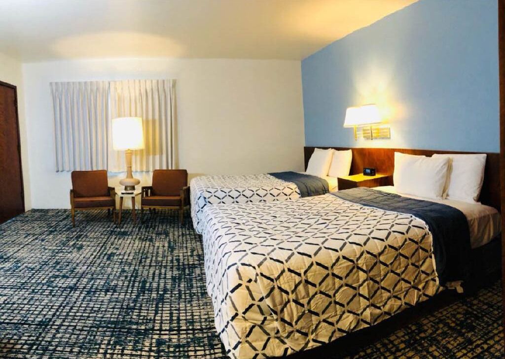 Quarto de hotel com duas camas, uma escrivaninha com cadeira, uma luminária e uma janela com cortinas.