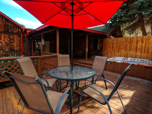 Um deck de madeira com uma mesa redonda e cadeiras, um guarda-sol vermelho e uma banheira de hidromassagem.