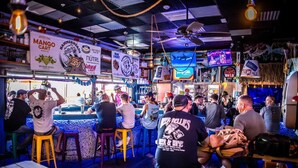 Um bar movimentado com banquetas e balcão, diversas marcas de cerveja expostas e um letreiro de neon ao fundo.