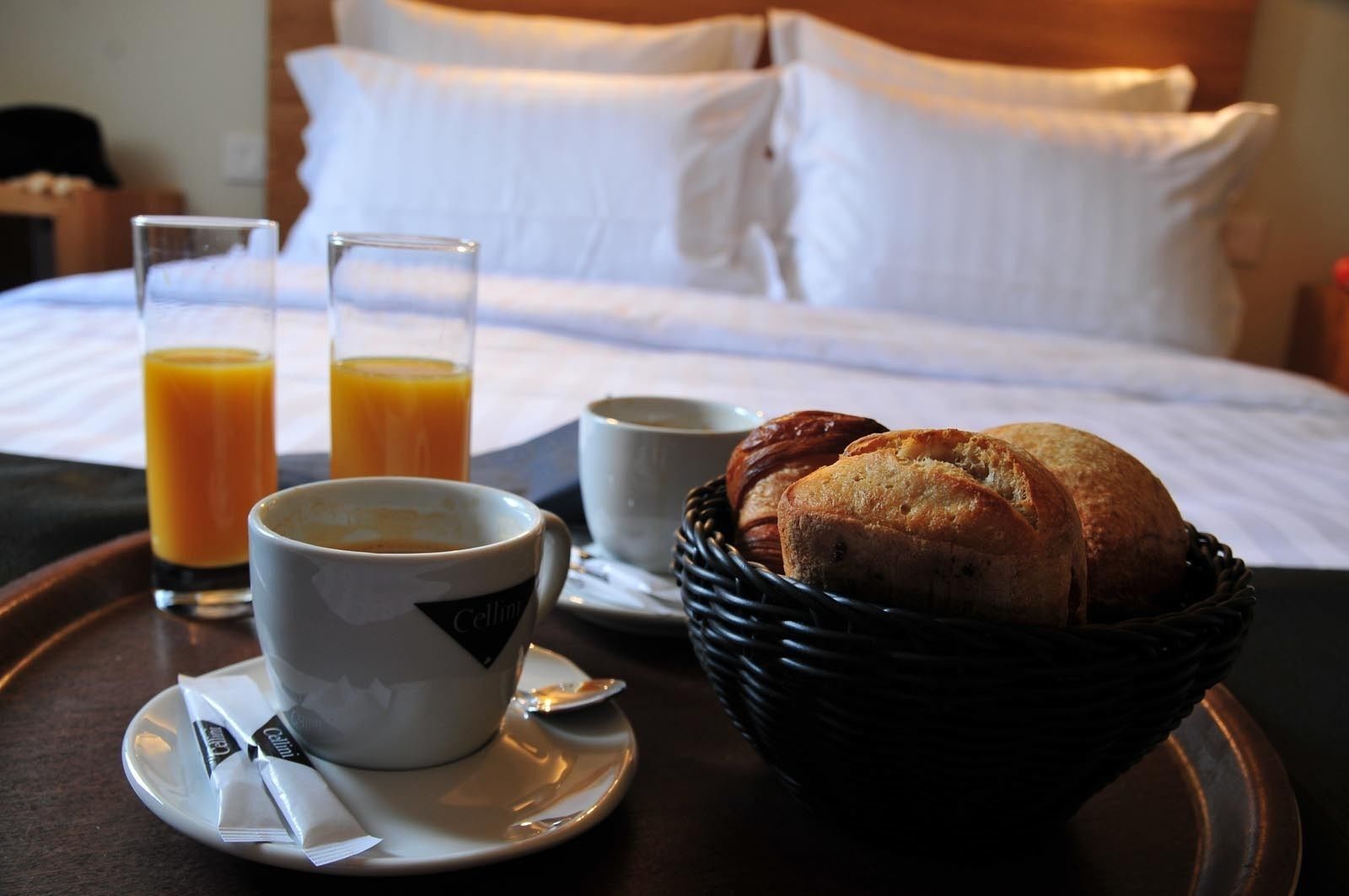 Bandeja de café da manhã com dois copos de suco de laranja, uma xícara de café, um prato com croissants e um guardanapo sobre uma cama com roupa de cama branca e almofadas.