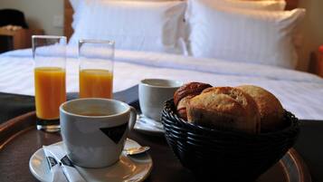 Bandeja de café da manhã com dois copos de suco de laranja, uma xícara de café, um prato com croissants e um guardanapo sobre uma cama com roupa de cama branca e almofadas.