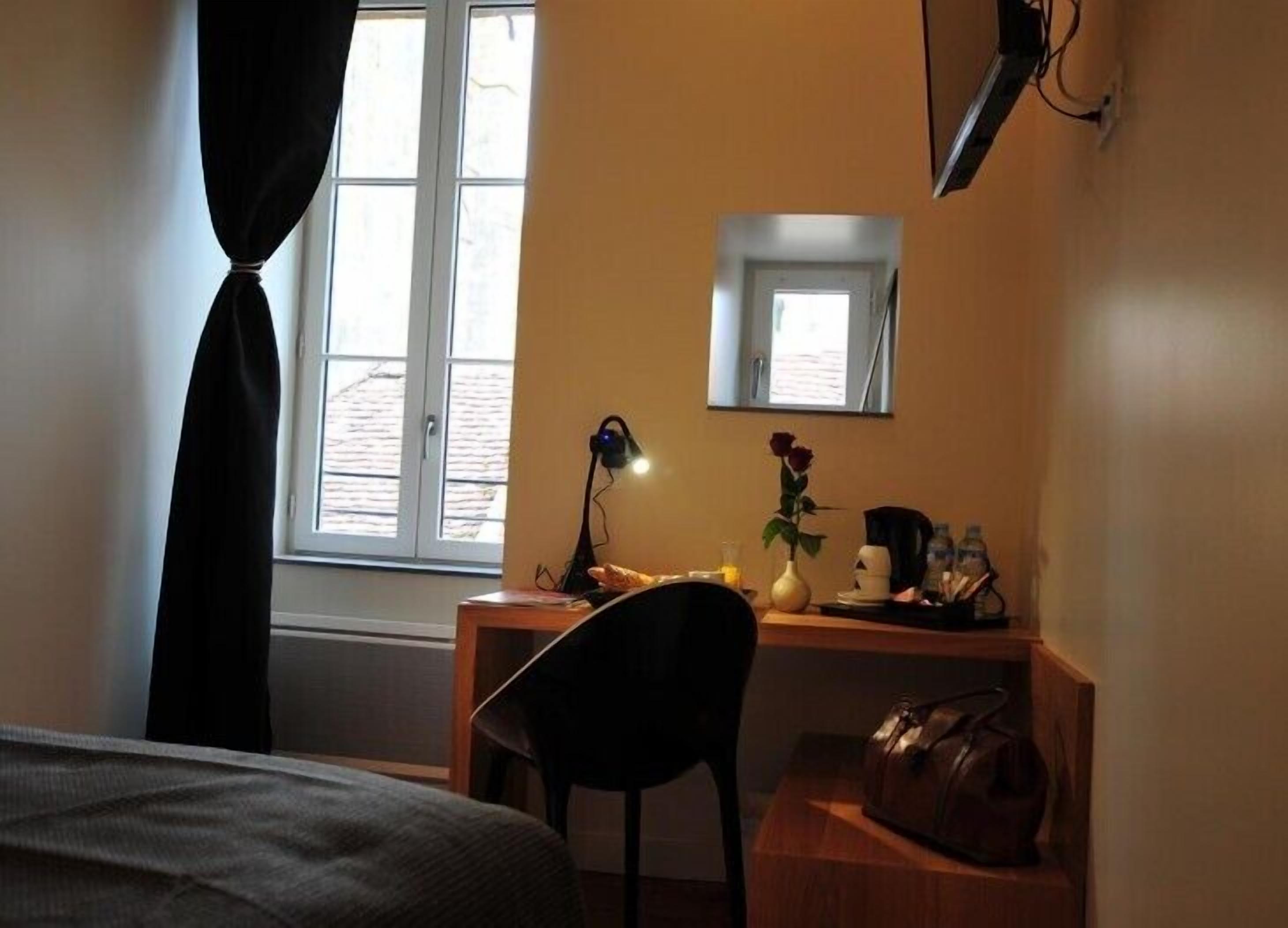 Quarto de hotel com cama, escrivaninha com cadeira, luminária de mesa, vaso com flor, cafeteira e bolsa sobre uma mesinha de cabeceira.