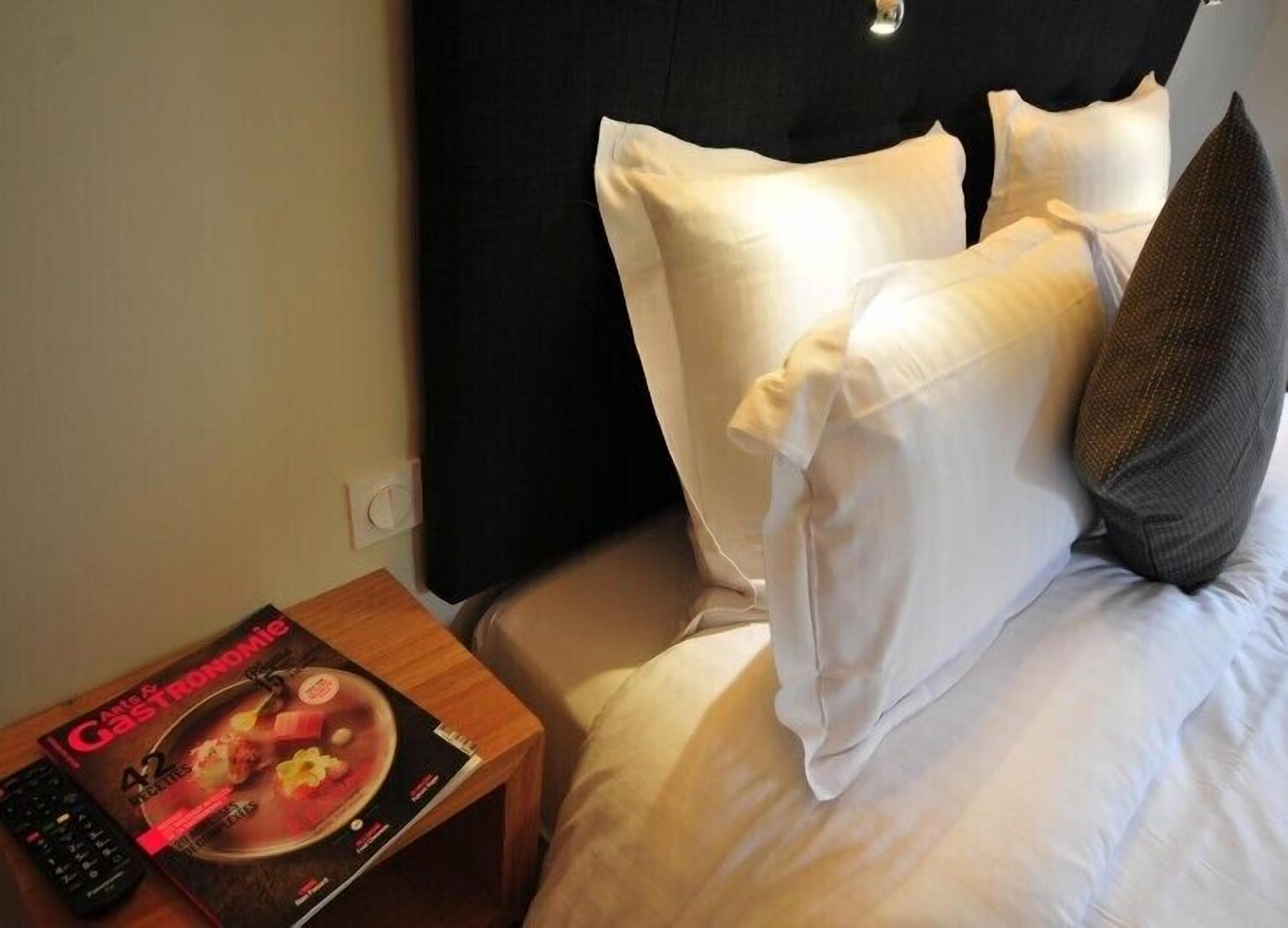 Uma cama bem arrumada com lençóis brancos e um travesseiro, uma mesa de cabeceira de madeira com uma revista, e uma cabeceira preta.
