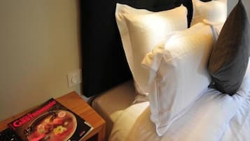 Uma cama bem arrumada com lençóis brancos e um travesseiro, uma mesa de cabeceira de madeira com uma revista, e uma cabeceira preta.