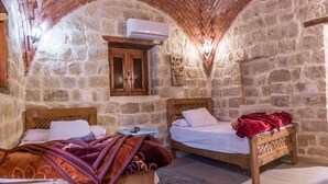 Quarto com duas camas de madeira, paredes de pedra, teto de tijolo e uma pequena janela.
