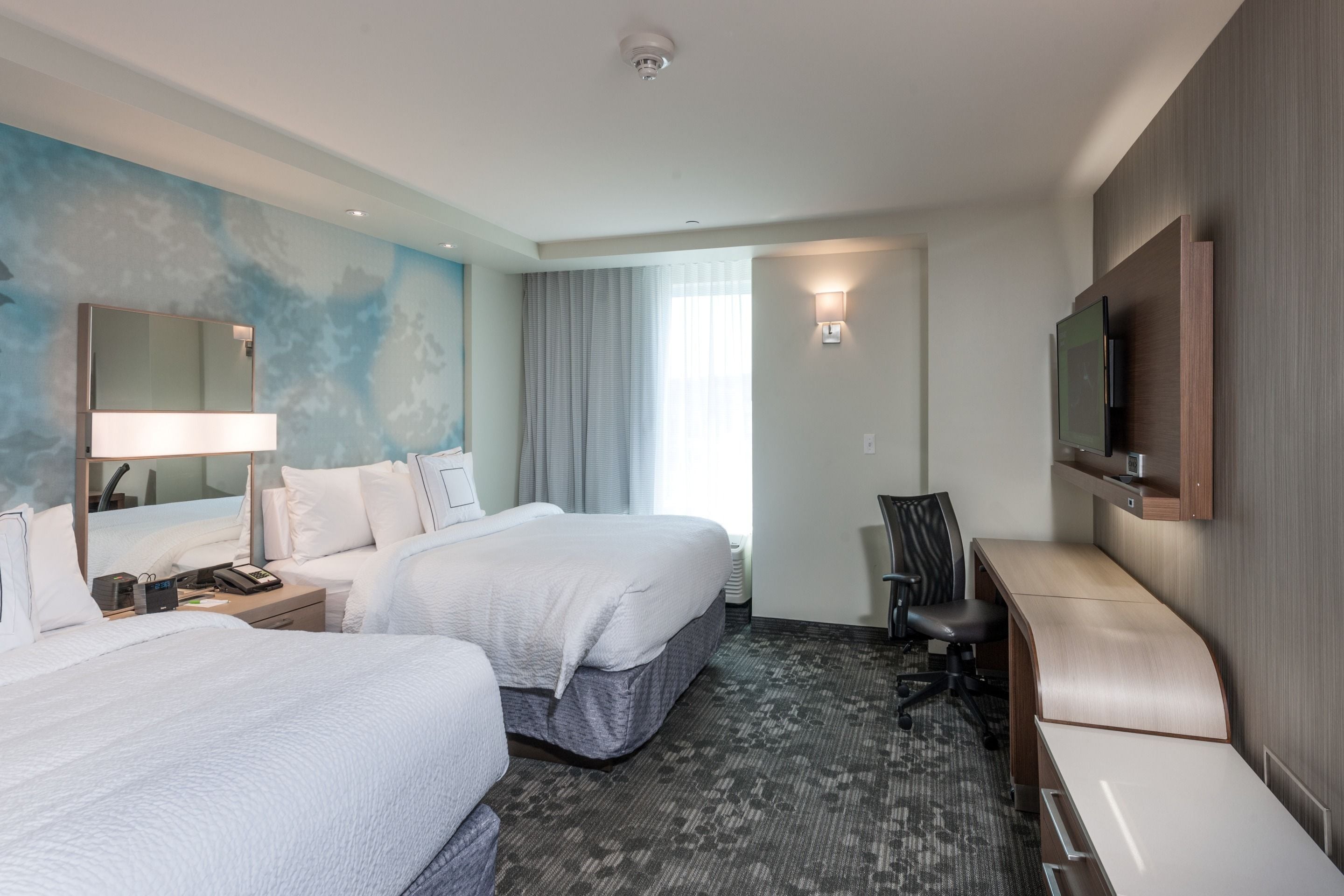 Quarto de hotel com duas camas, uma escrivaninha, uma cadeira, uma televisão e uma janela grande com cortinas.