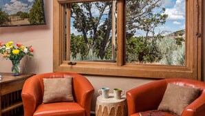 Um quarto com duas poltronas de couro, uma mesa de madeira pequena com um vaso de flores, e uma janela grande com vista para árvores e céu.