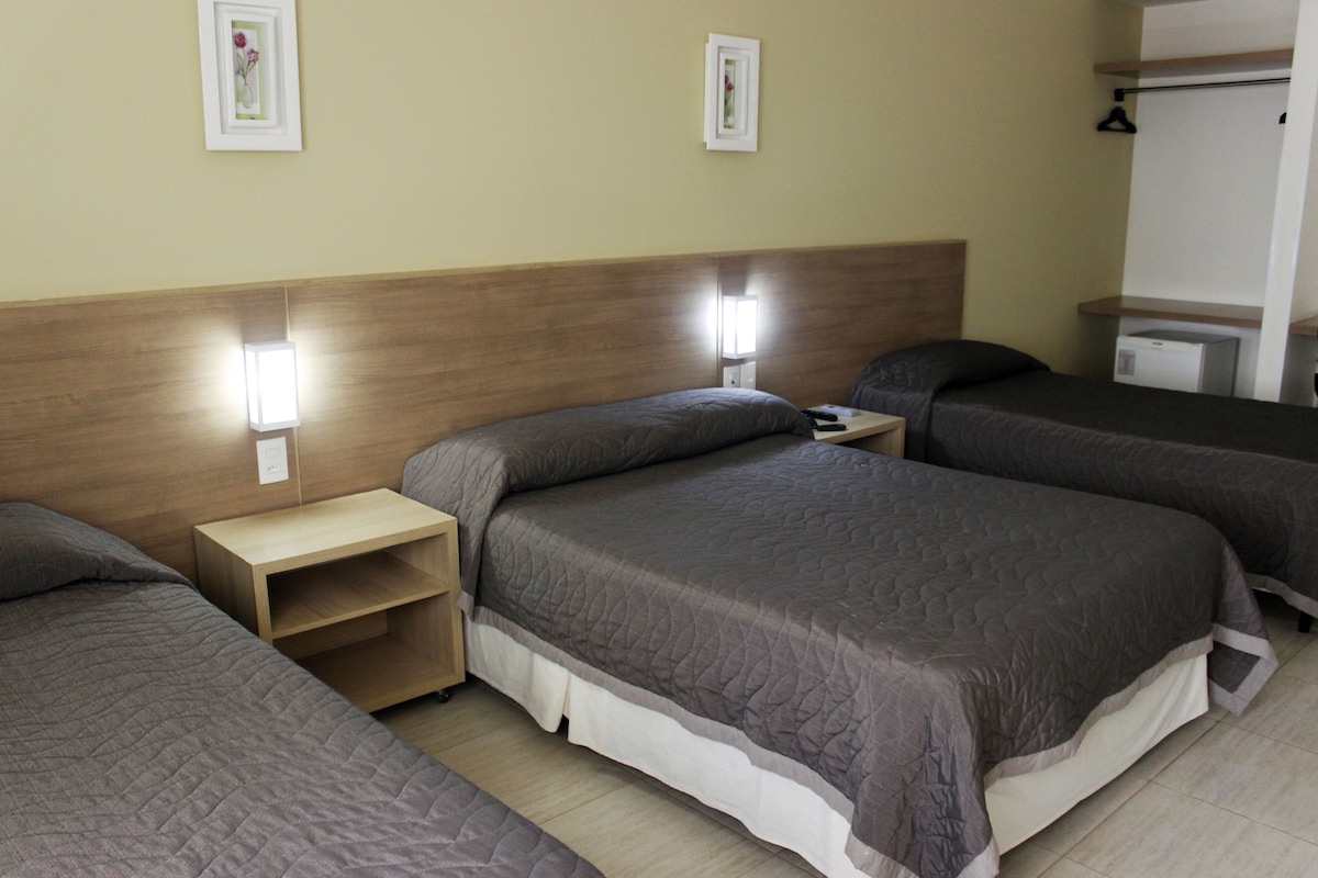 Quarto de hotel com duas camas, uma mesinha de cabeceira, um criado-mudo e uma prateleira na parede.
