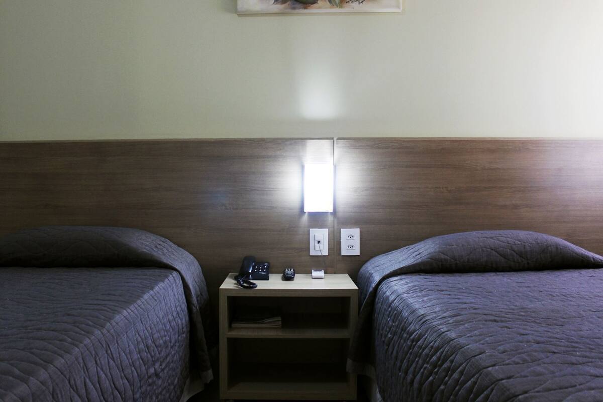 Quarto de hotel com duas camas de solteiro, uma mesinha de cabeceira com telefone e tomada, e uma luminária fixada na parede.
