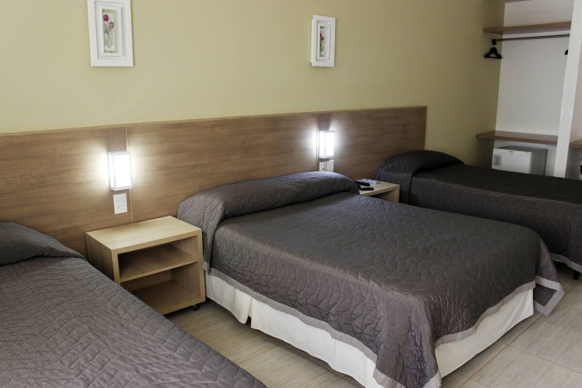 Quarto de hotel com duas camas, uma mesinha de cabeceira, um criado-mudo e uma prateleira na parede.
