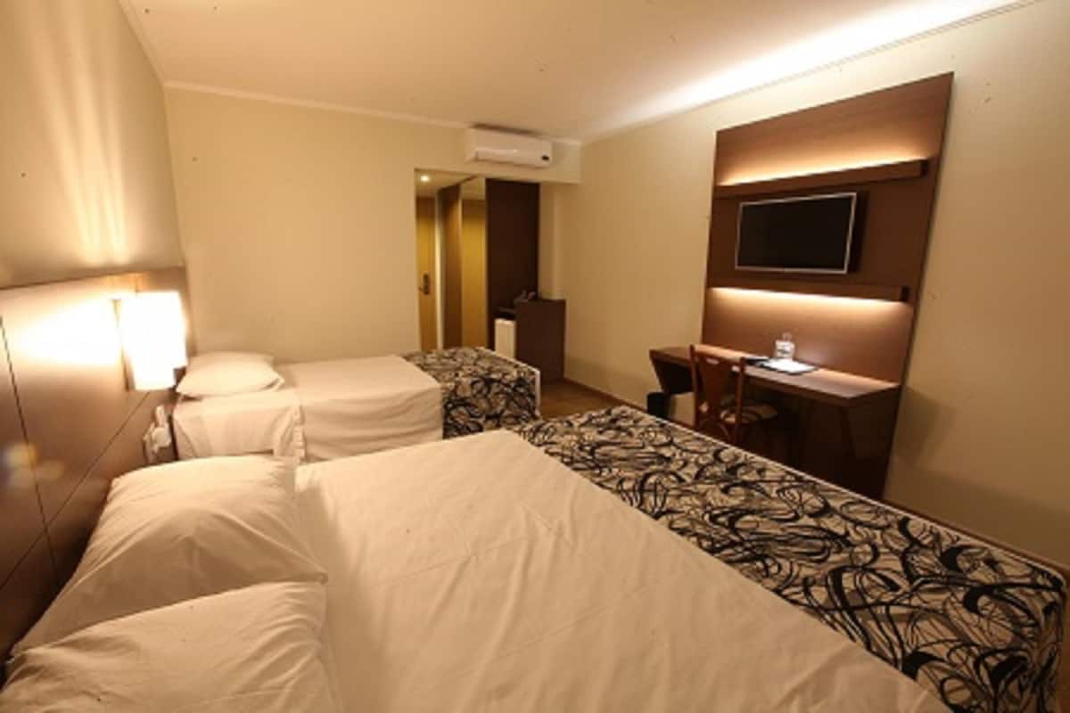Quarto de hotel com duas camas, uma escrivaninha, uma cadeira, uma televisão e ar condicionado.