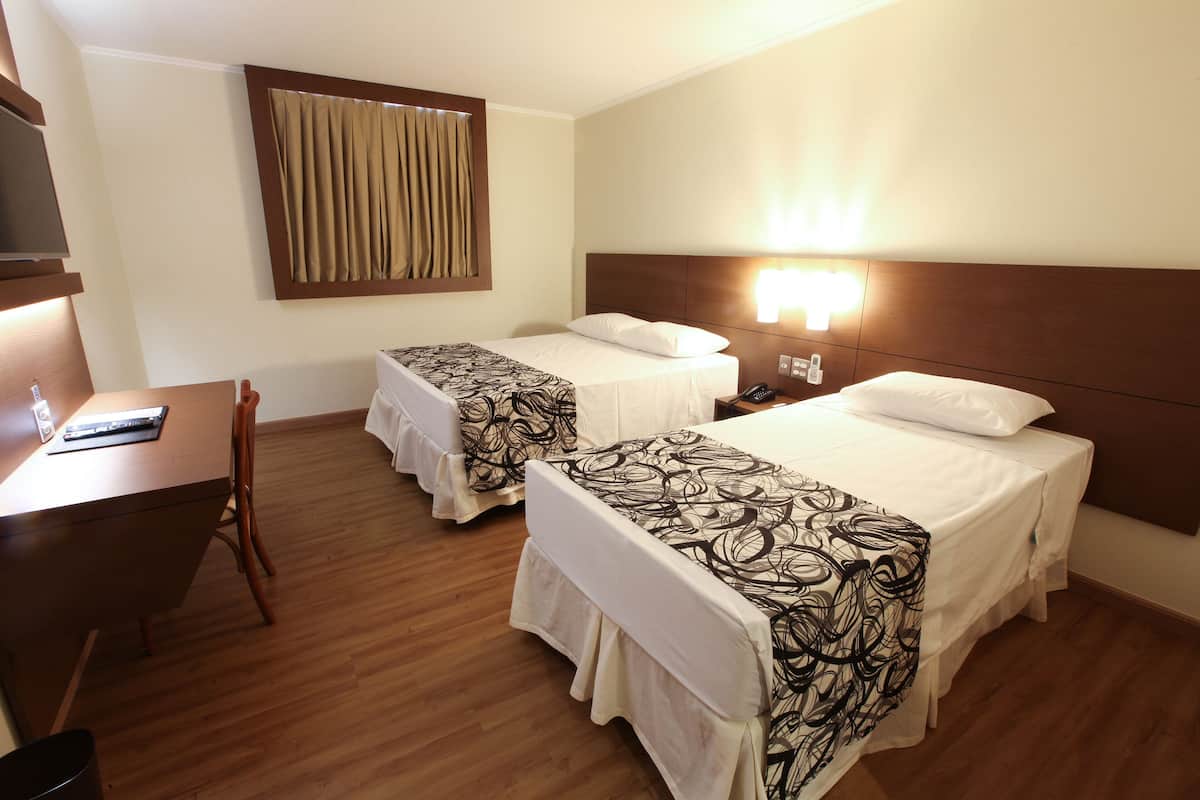 Quarto de hotel com duas camas, uma escrivaninha, uma cadeira e uma televisão.