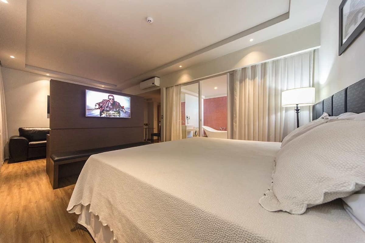 Quarto de hotel moderno com uma cama grande, televisão de tela plana na parede, área de estar com sofá e uma janela ampla com cortinas.