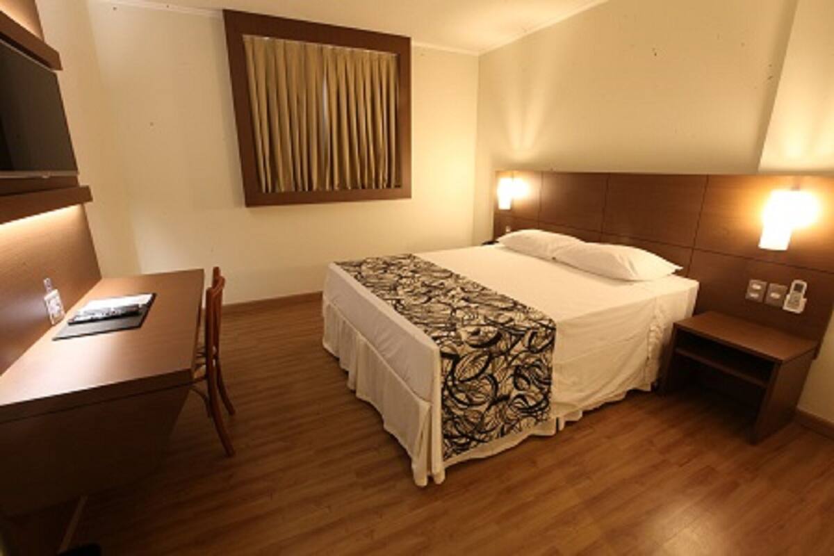 Quarto de hotel com cama, mesa de cabeceira, escrivaninha e cadeira.