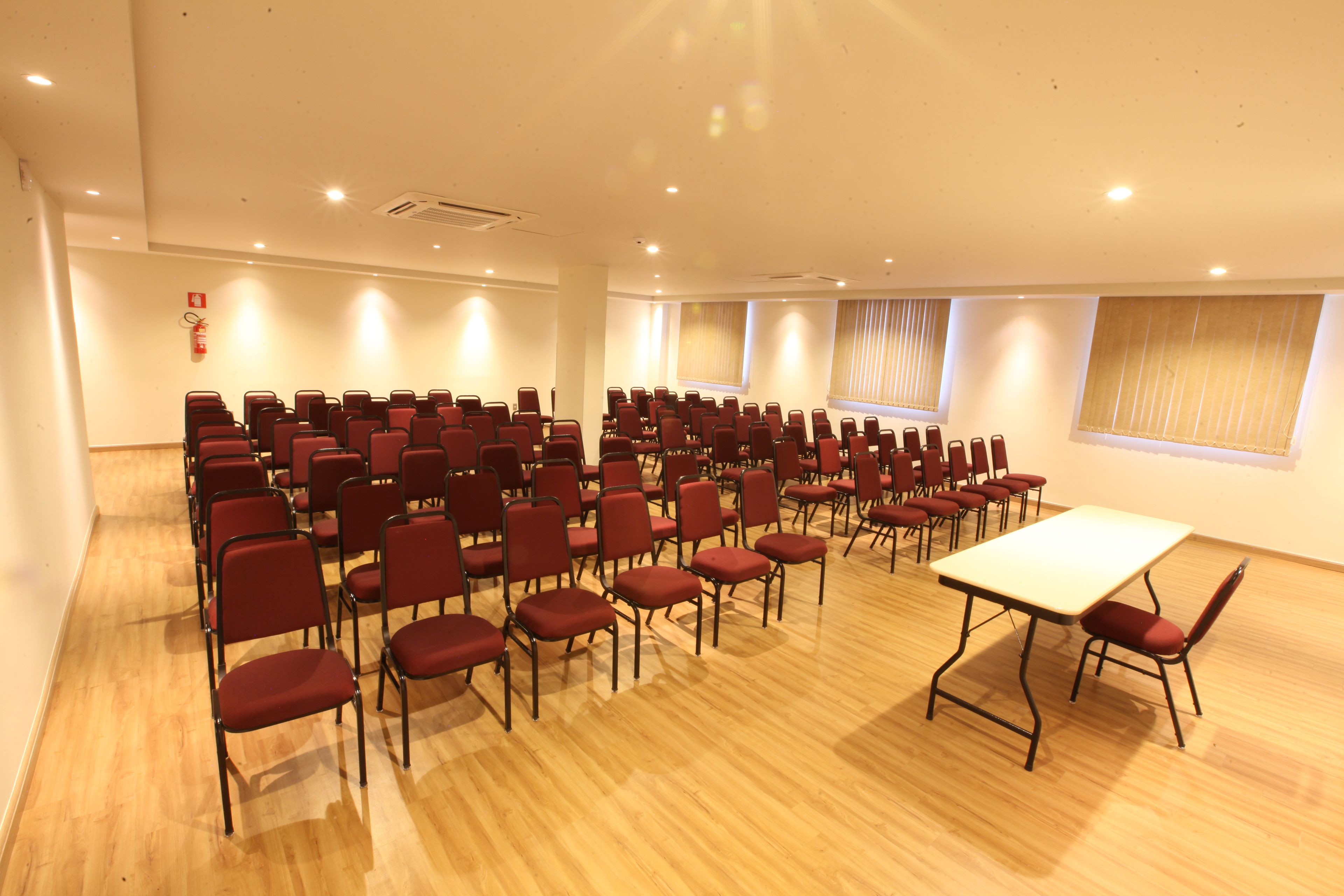 Uma sala de conferências com fileiras de cadeiras vermelhas, uma mesa branca e janelas amplas com persianas.