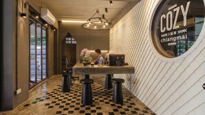 Um saguão moderno de hotel com piso xadrez, balcão de recepção e uma grande janela circular com a inscrição 'COZY Chiangmai'.