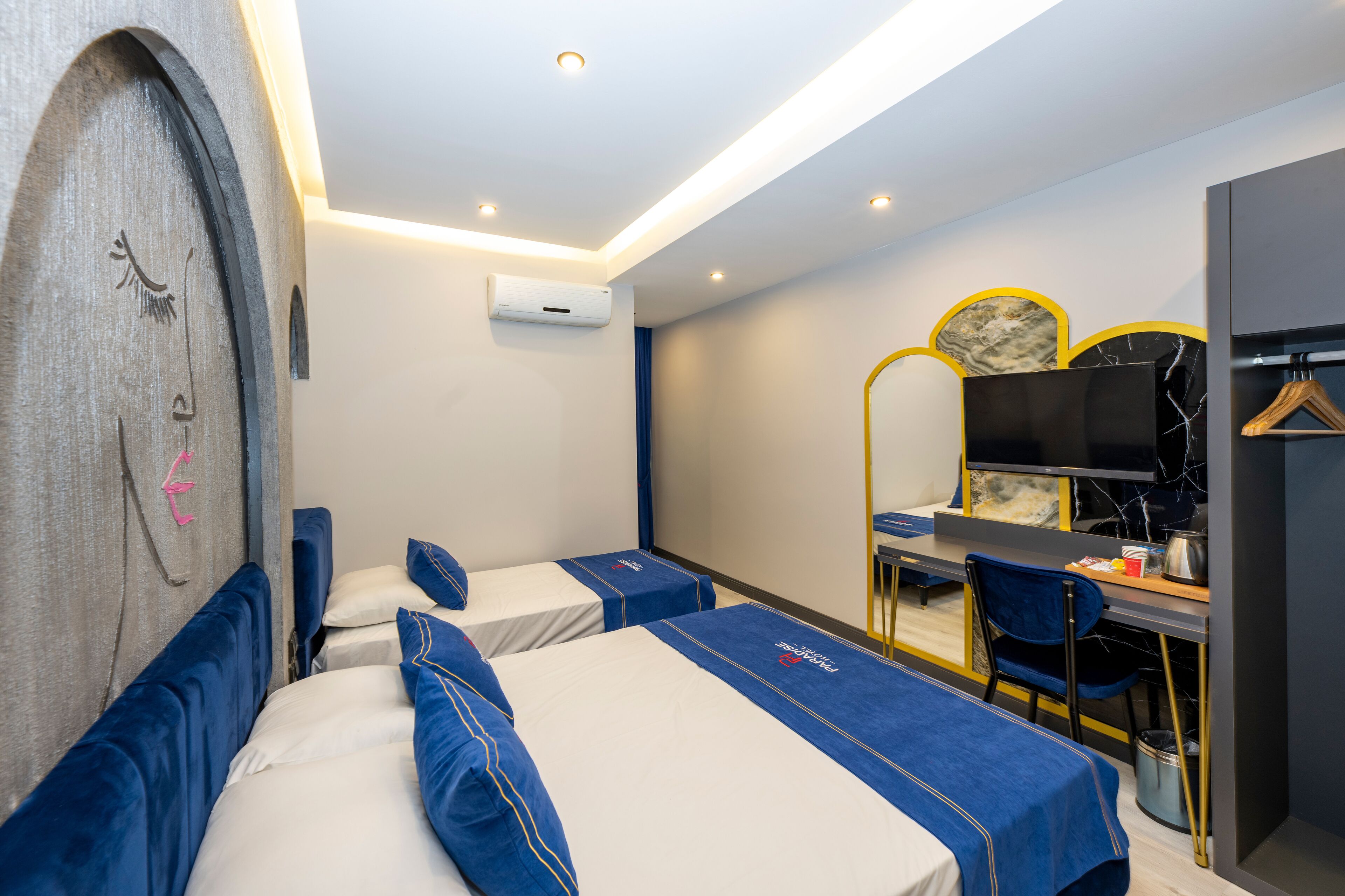 Quarto de hotel com duas camas, uma escrivaninha, uma cadeira, uma televisão e um guarda-roupa.