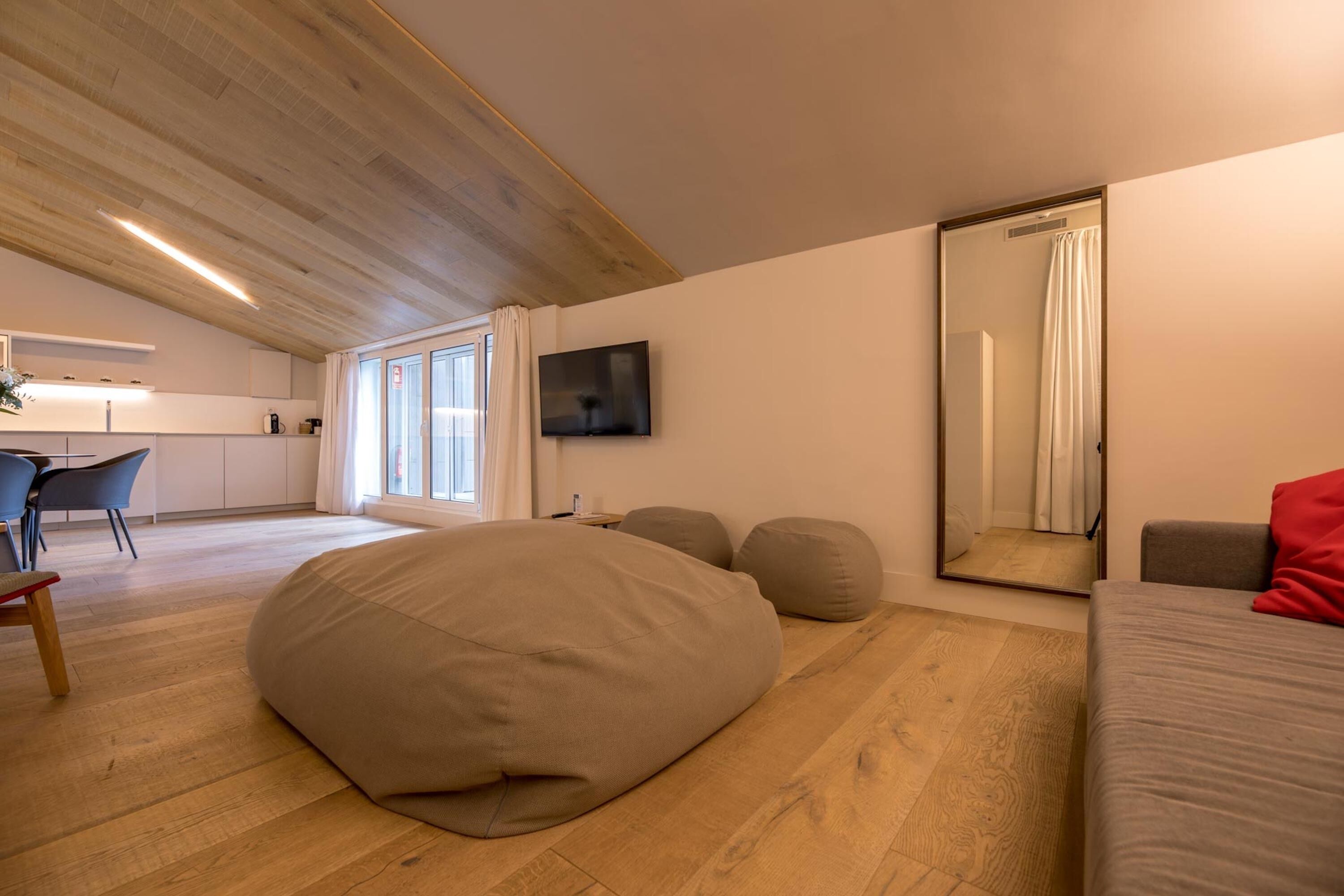 Quarto moderno com teto de madeira, um grande puff, sofá com almofada vermelha, televisão e espelho.