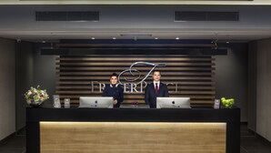 Uma receção com dois funcionários, um painel de ripas de madeira com a palavra "RECEPTION" ao fundo, e um design moderno com bancada preta e dois computadores portáteis.