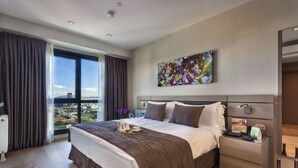 Quarto de hotel moderno com uma cama grande, mesa de cabeceira, pintura e vista para a cidade.