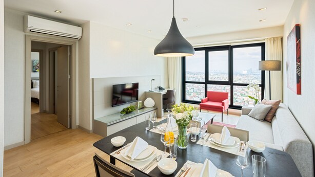 Quarto de hotel moderno com área de refeições, televisão, sofá e uma grande janela com vista para a cidade.