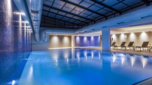 Piscina interior moderna com água azul cristalina, espreguiçadeiras e teto alto com dutos aparentes.