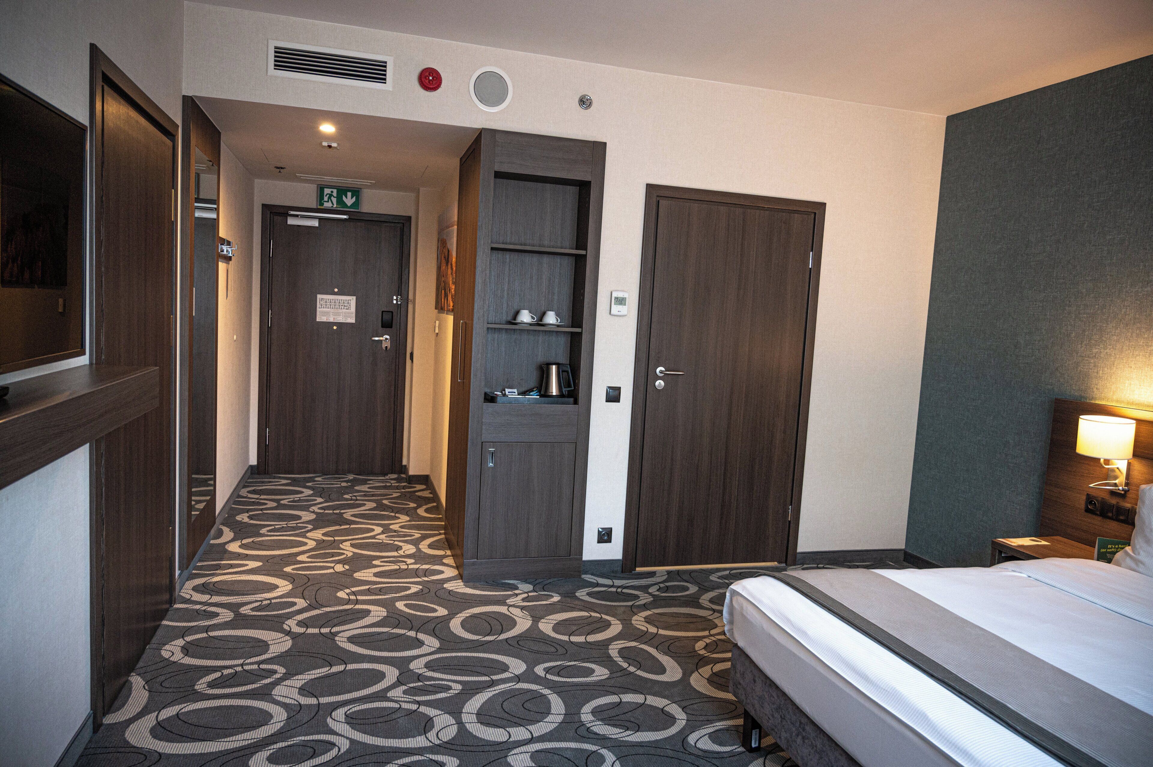 Quarto de hotel com carpete estampado, uma cama, uma mesa de cabeceira, uma luminária na parede e uma porta para outro quarto.