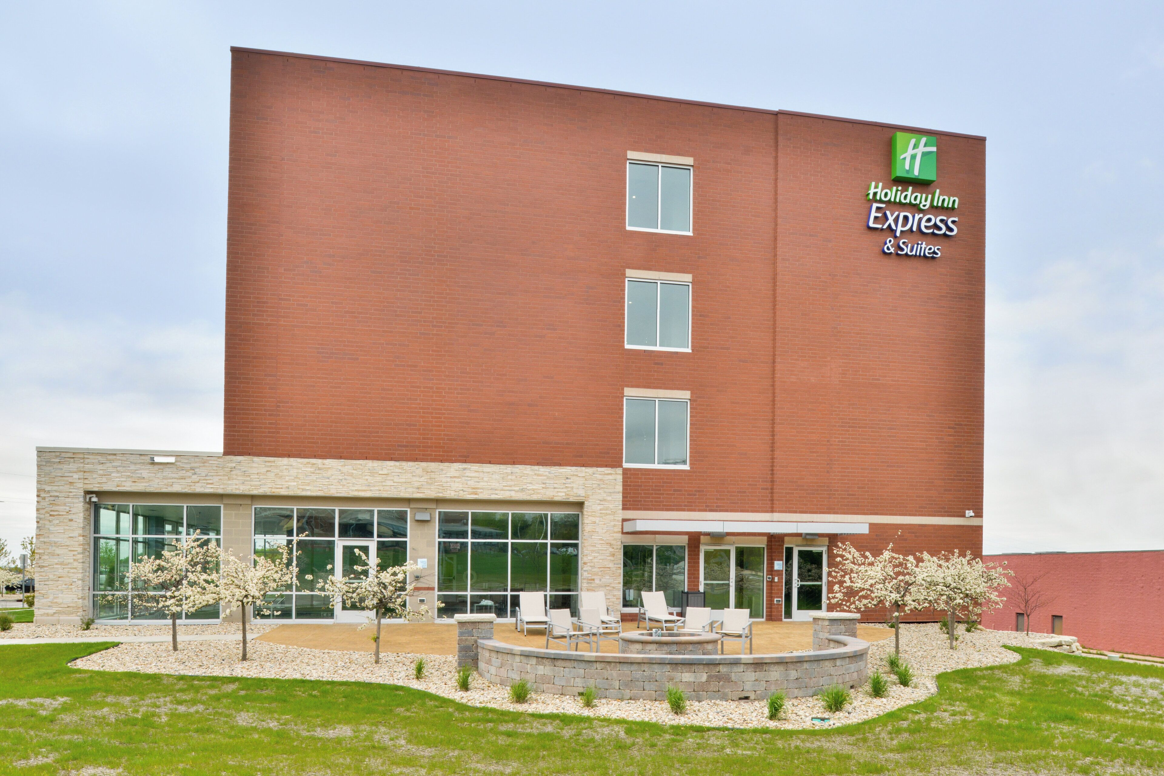 Edifício Holiday Inn Express & Suites com fachada em tijolos, amplas janelas e área externa ajardinada com mobiliário.