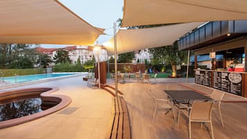 Um pátio externo com piscina, deck de madeira e área de bar com cardápios.