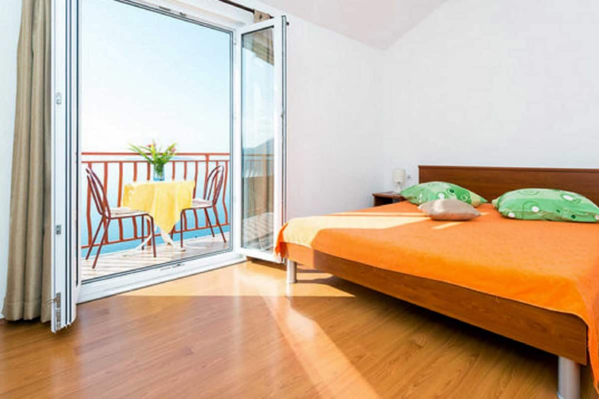 Quarto com cama de madeira, mesa de cabeceira e vista para uma varanda com mesa e cadeiras.