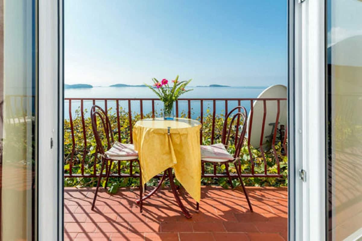 Uma varanda com mesa e cadeiras, um vaso com flores e vista para o mar.