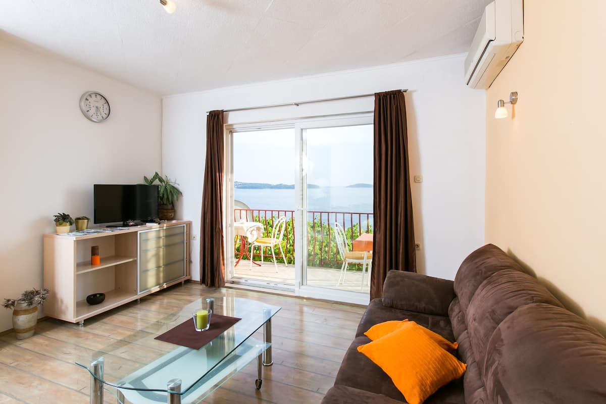 Uma sala de estar com mesa de centro de vidro, sofá marrom com almofada laranja, televisão sobre um móvel de madeira e varanda com vista para o mar.