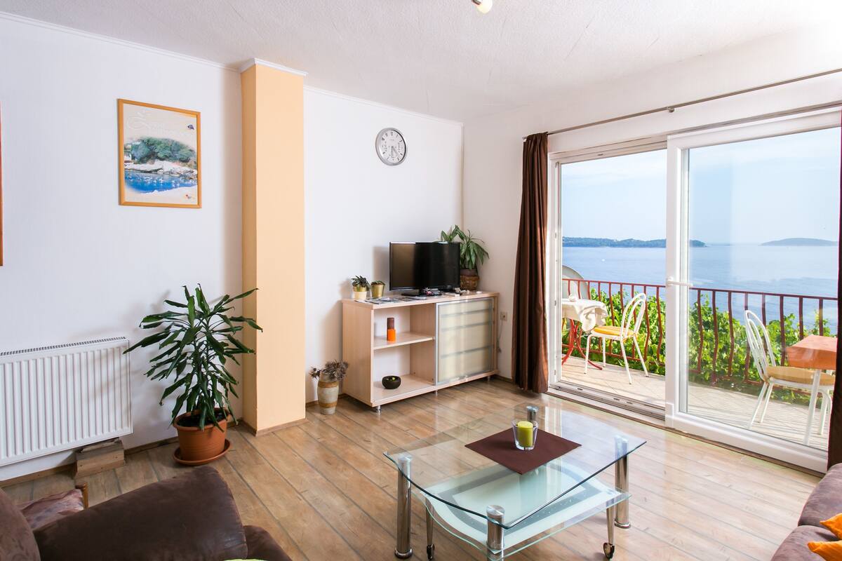 Uma sala de estar moderna com uma mesa de centro de vidro, um sofá, uma TV em um móvel e vista para o mar através de grandes janelas.