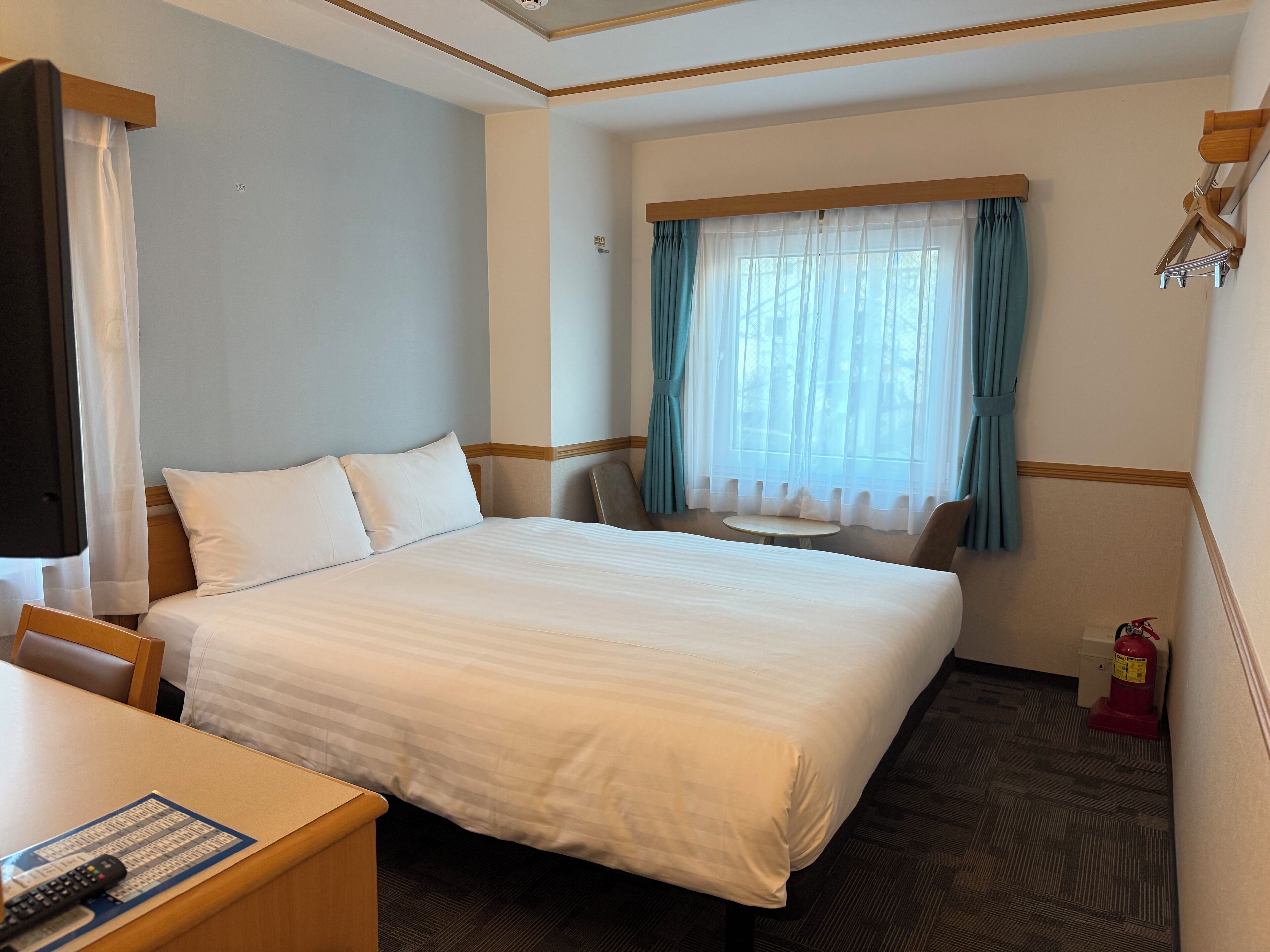 Quarto de hotel com uma cama grande, uma mesinha, uma cadeira, televisão, janela com cortinas e extintor de incêndio.
