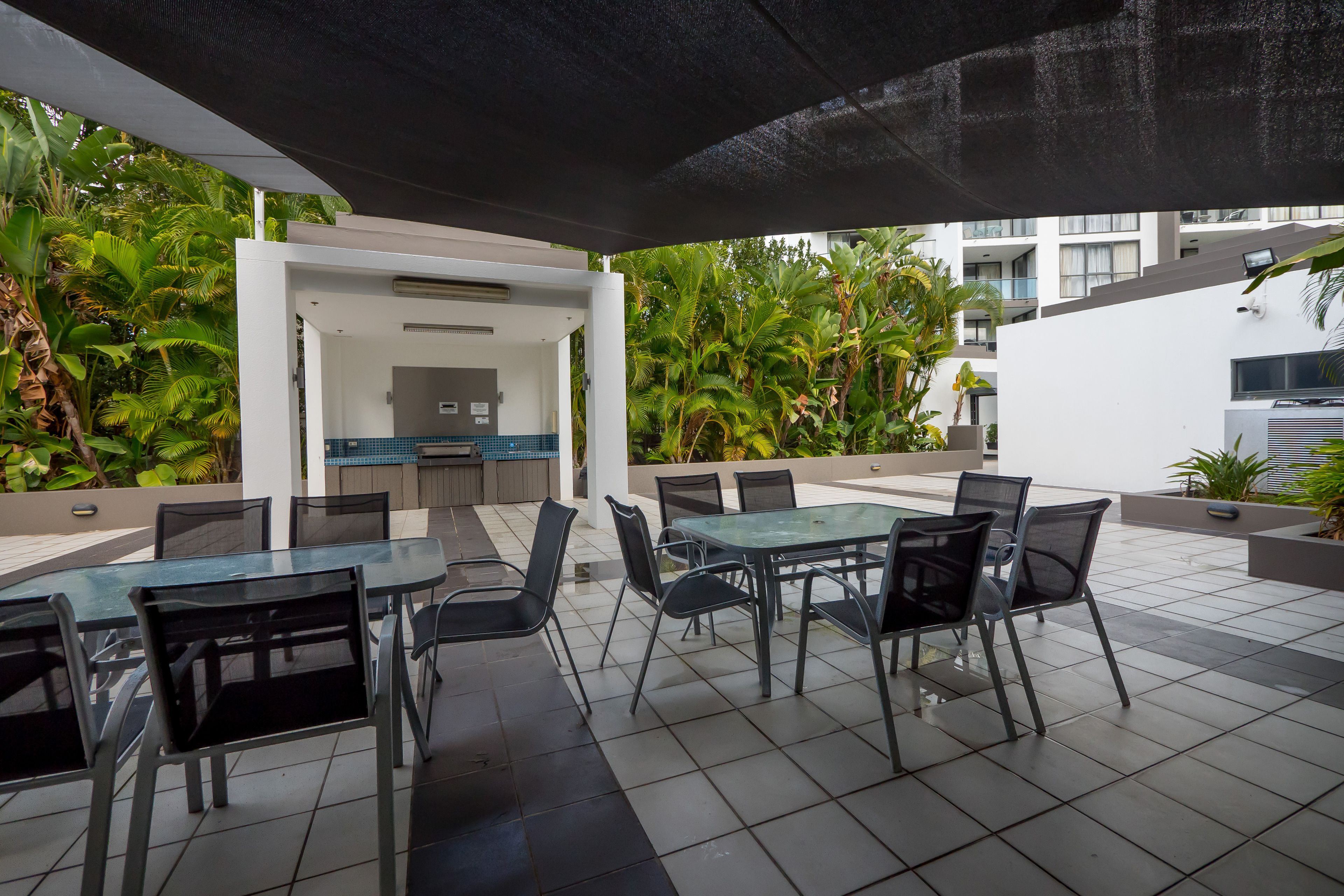 Um pátio externo com mesa e cadeiras de vidro, cercado por vegetação exuberante e um edifício branco ao fundo.