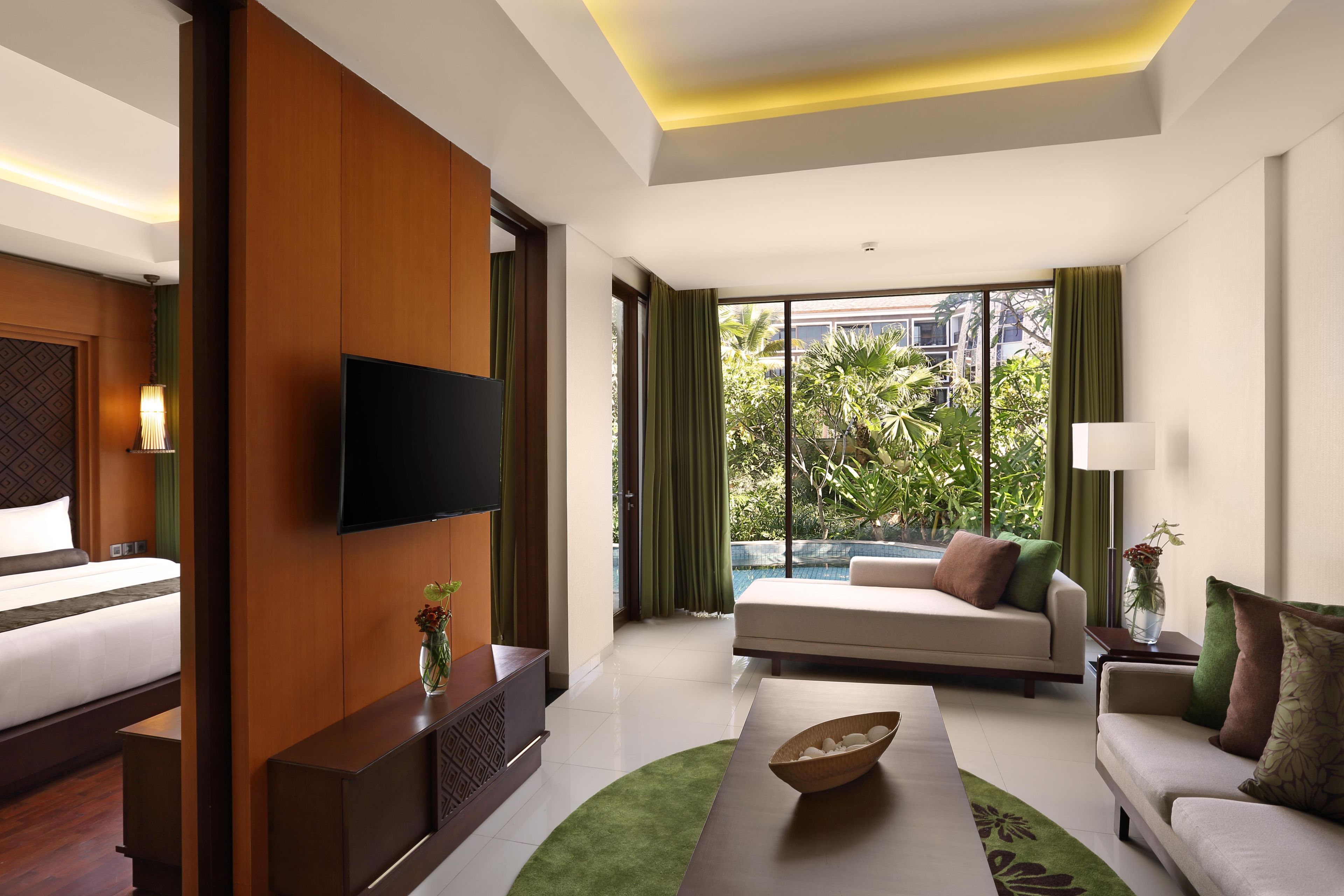 Quarto de hotel moderno com cama, televisão de tela plana, sofá, mesa de centro e vista para uma piscina e vegetação.