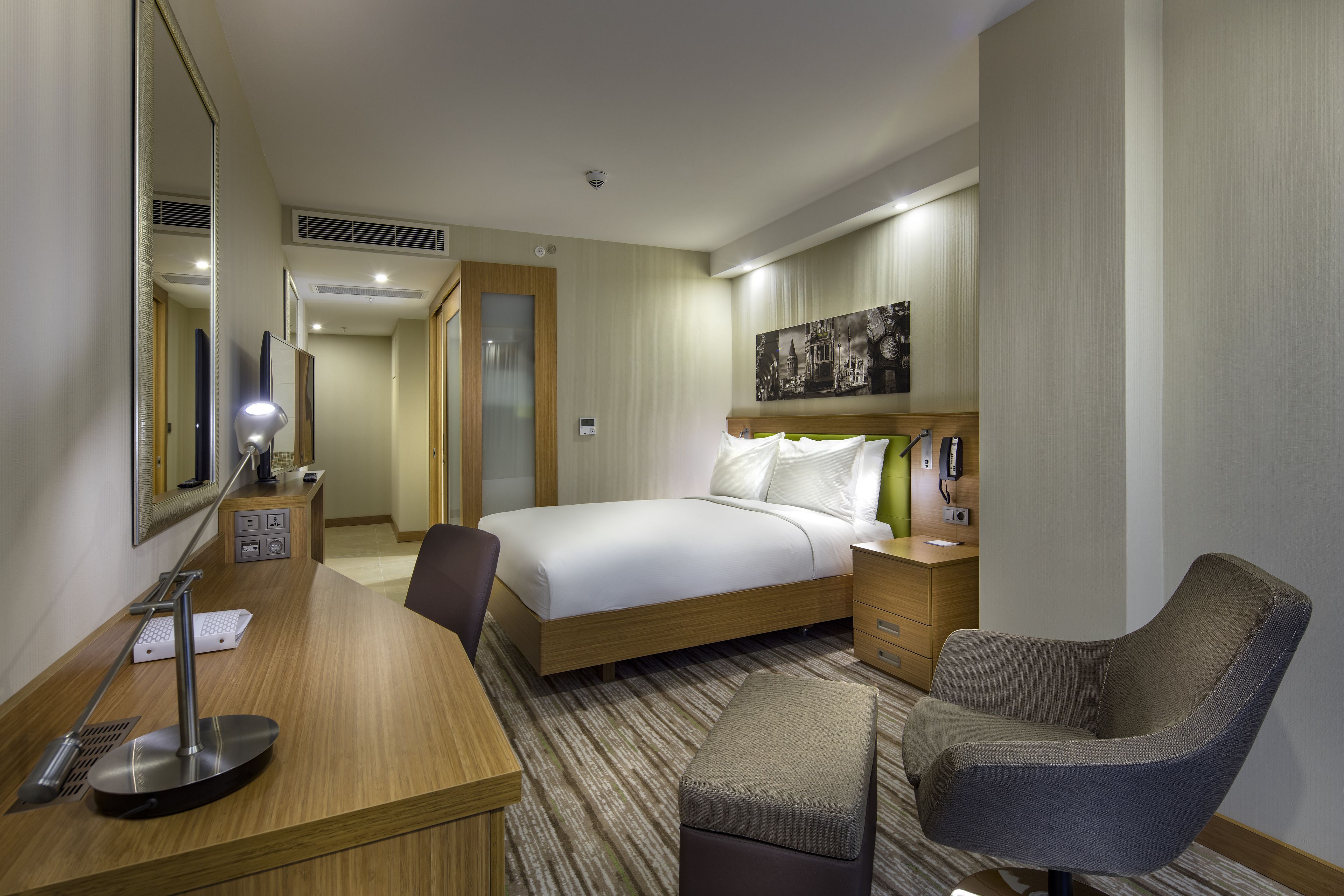 Quarto de hotel com cama, escrivaninha com luminária, cadeira, mesa de cabeceira e um quadro na parede.
