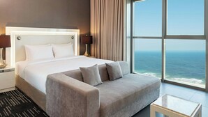 Quarto de hotel com cama, sofá, mesa de cabeceira, luminária, janela com vista para o oceano e uma pequena mesa.