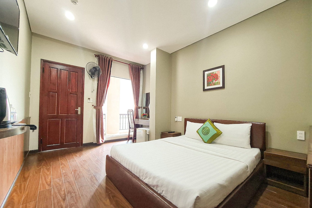 Quarto de hotel com cama, piso de madeira, escrivaninha com cadeira, ventoinha e um quadro na parede.