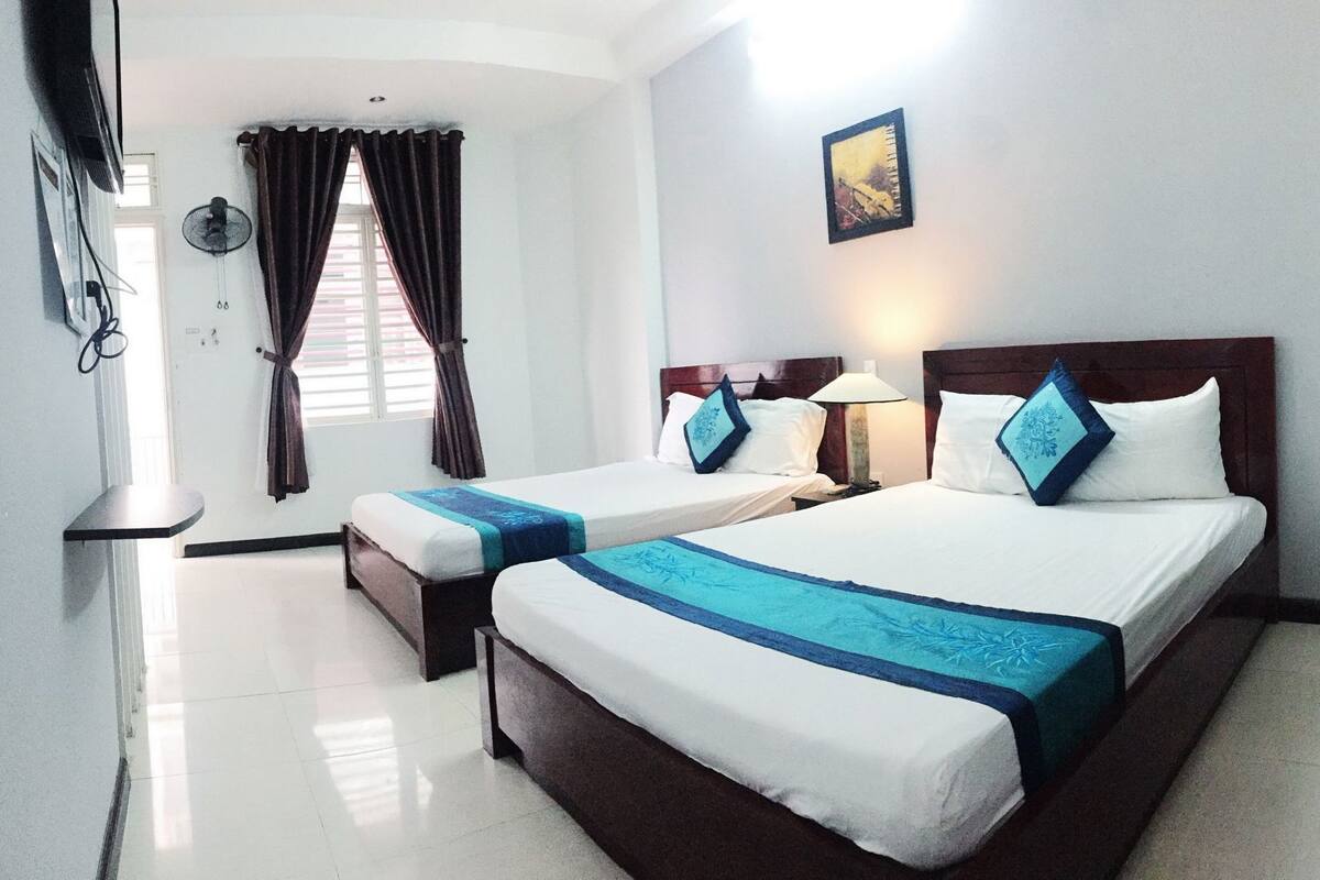Quarto de hotel com duas camas, uma mesa de cabeceira com abajur, um quadro na parede e uma janela com cortinas.