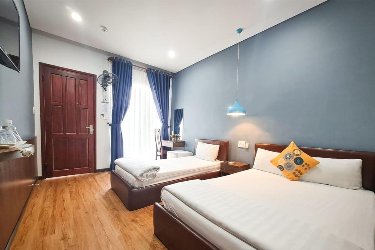 Quarto de hotel com duas camas, chão de madeira, parede azul, porta de madeira e luminária pendente azul.
