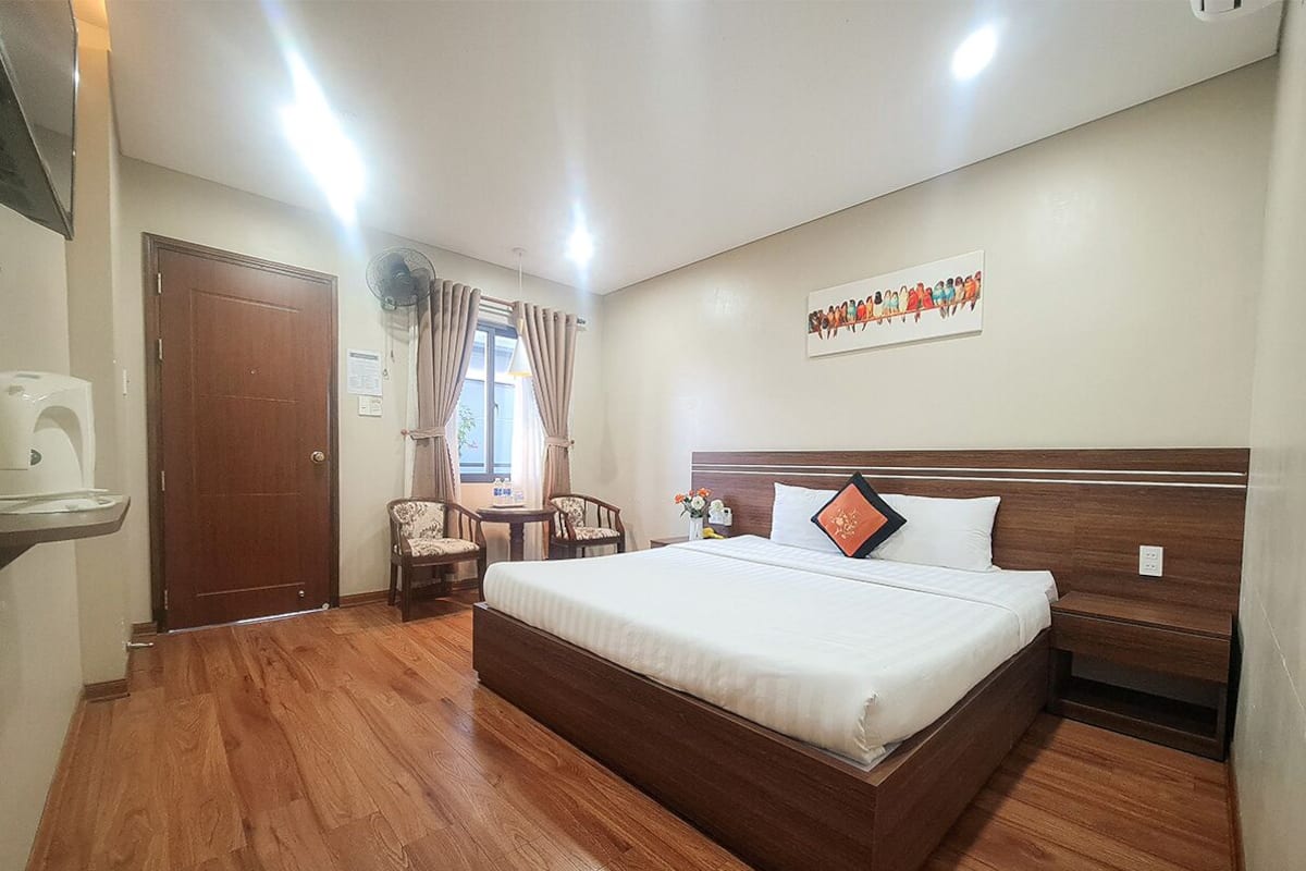 Quarto de hotel com piso de madeira, cama com roupa de cama branca, cabeceira de madeira, mesa de cabeceira, uma cadeira, escrivaninha, janela com cortinas e uma obra de arte na parede.