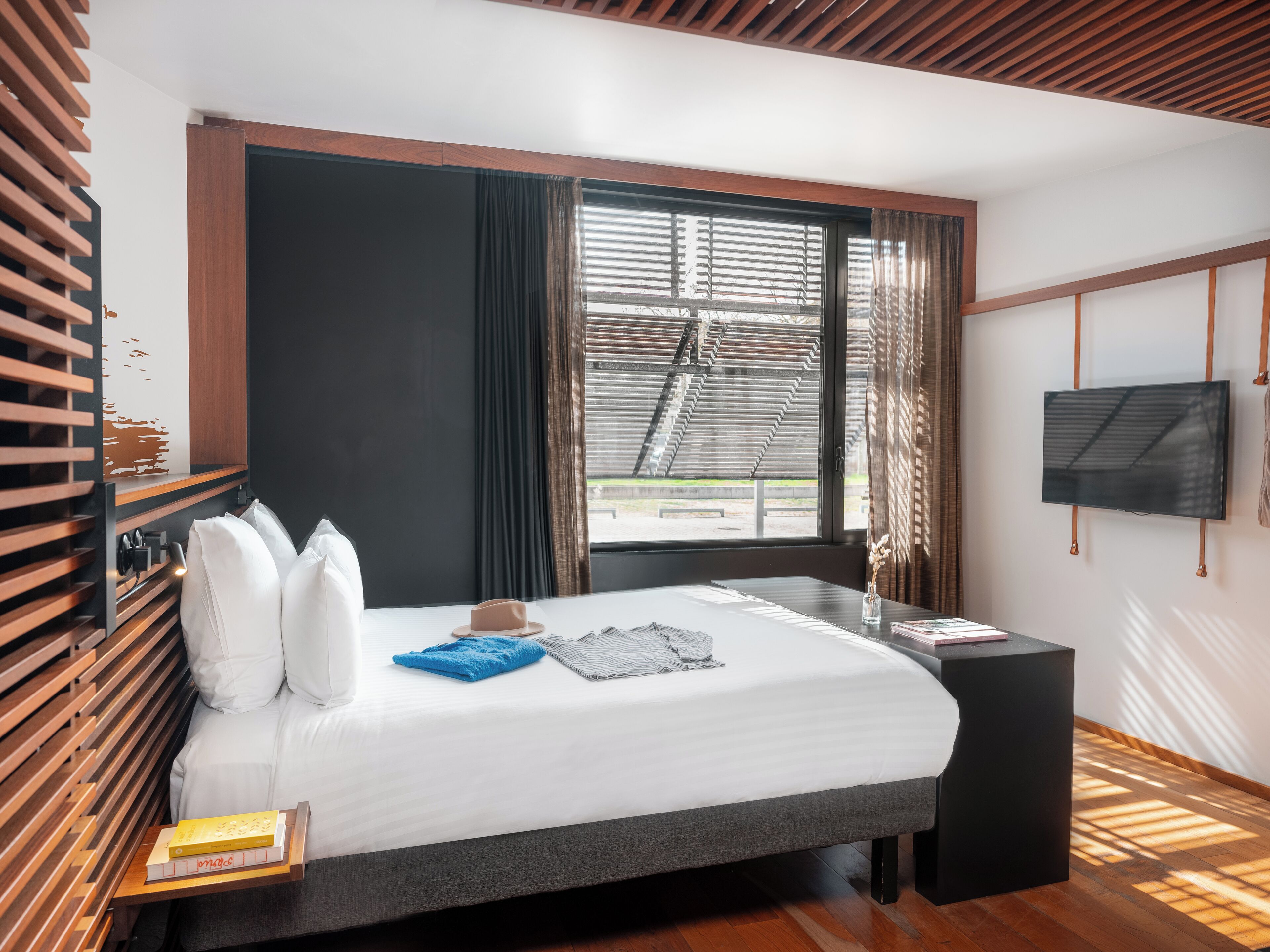 Quarto de hotel moderno com uma cama grande, televisão de tela plana na parede e uma janela com persianas.